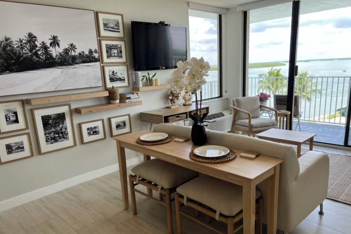 Uma sala de estar moderna com uma mesa de jantar de madeira posta para dois, um sofá e uma TV fixada na parede.