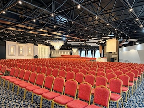 Um amplo espaço interno com fileiras de cadeiras vermelhas, um palco e teto alto com luminárias.