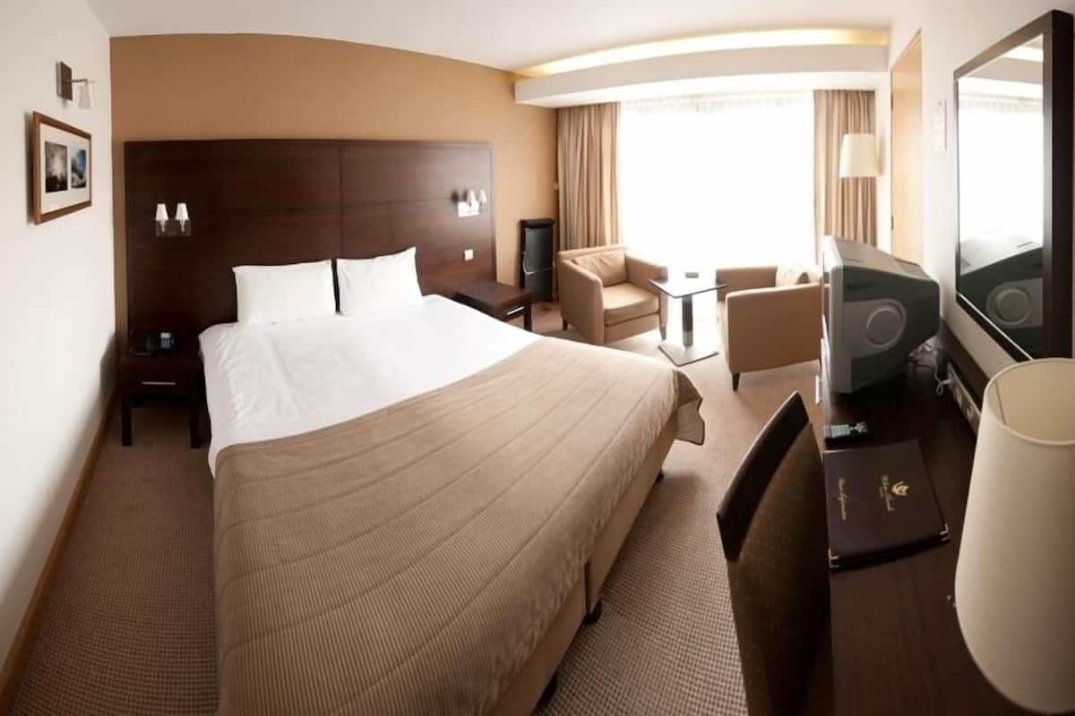 Quarto de hotel com uma cama grande, uma escrivaninha com cadeira, televisão e abajur.