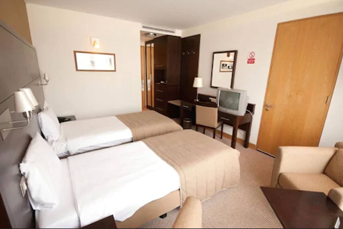 Quarto de hotel com duas camas, uma escrivaninha, uma cadeira, uma televisão e um guarda-roupa.