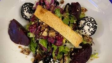 Um prato com salada de folhas verdes, folhas roxas e objetos redondos pretos e brancos, possivelmente azeitonas, com um pedaço de pão por cima.