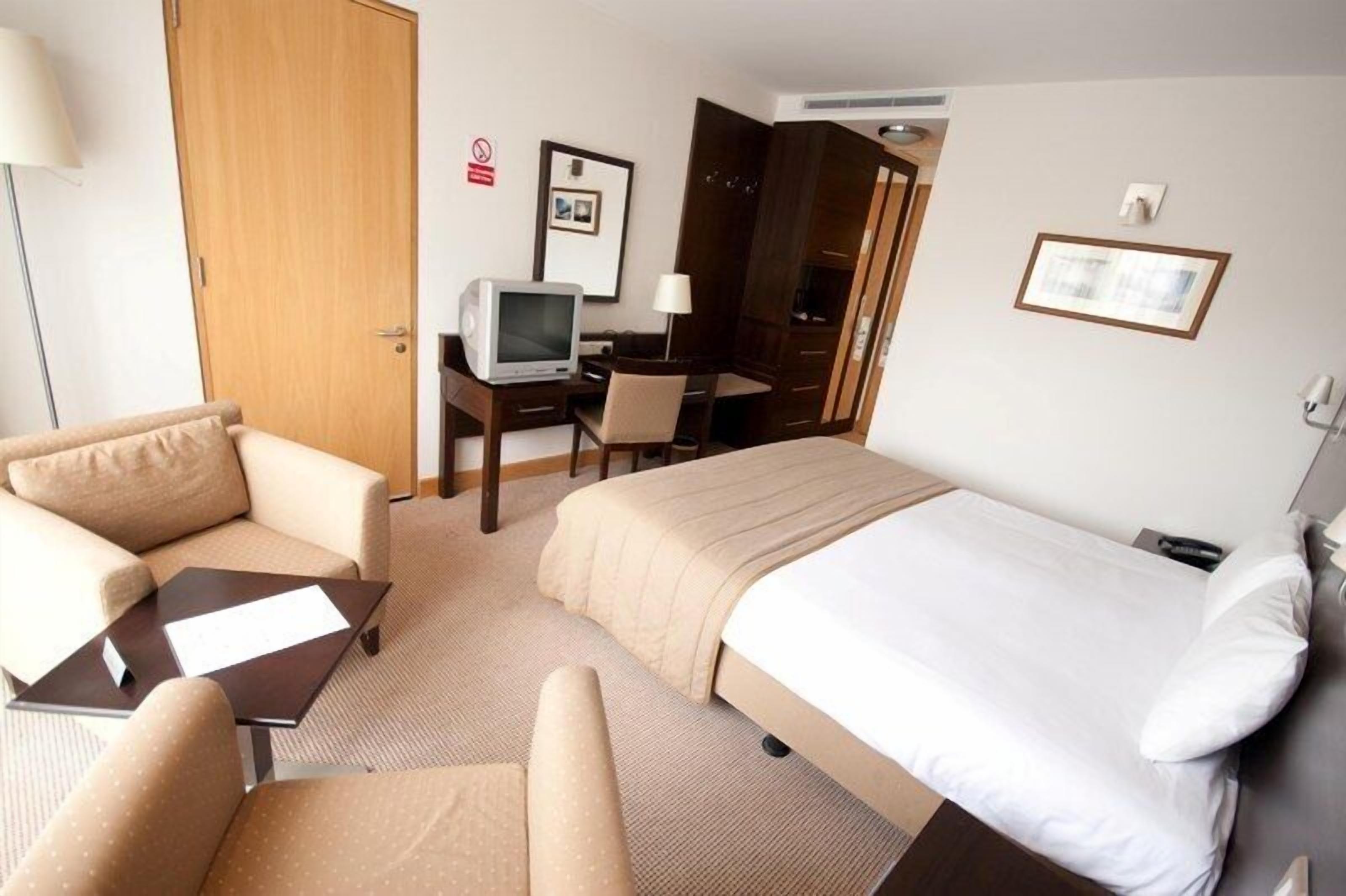 Quarto de hotel com cama, escrivaninha com computador, cadeira, sofá e abajur.