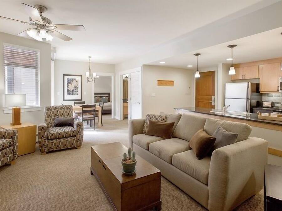 Uma sala de estar moderna com sofá, poltronas e mesa de centro. Há uma cozinha equipada com frigorífico, micro-ondas e fogão.