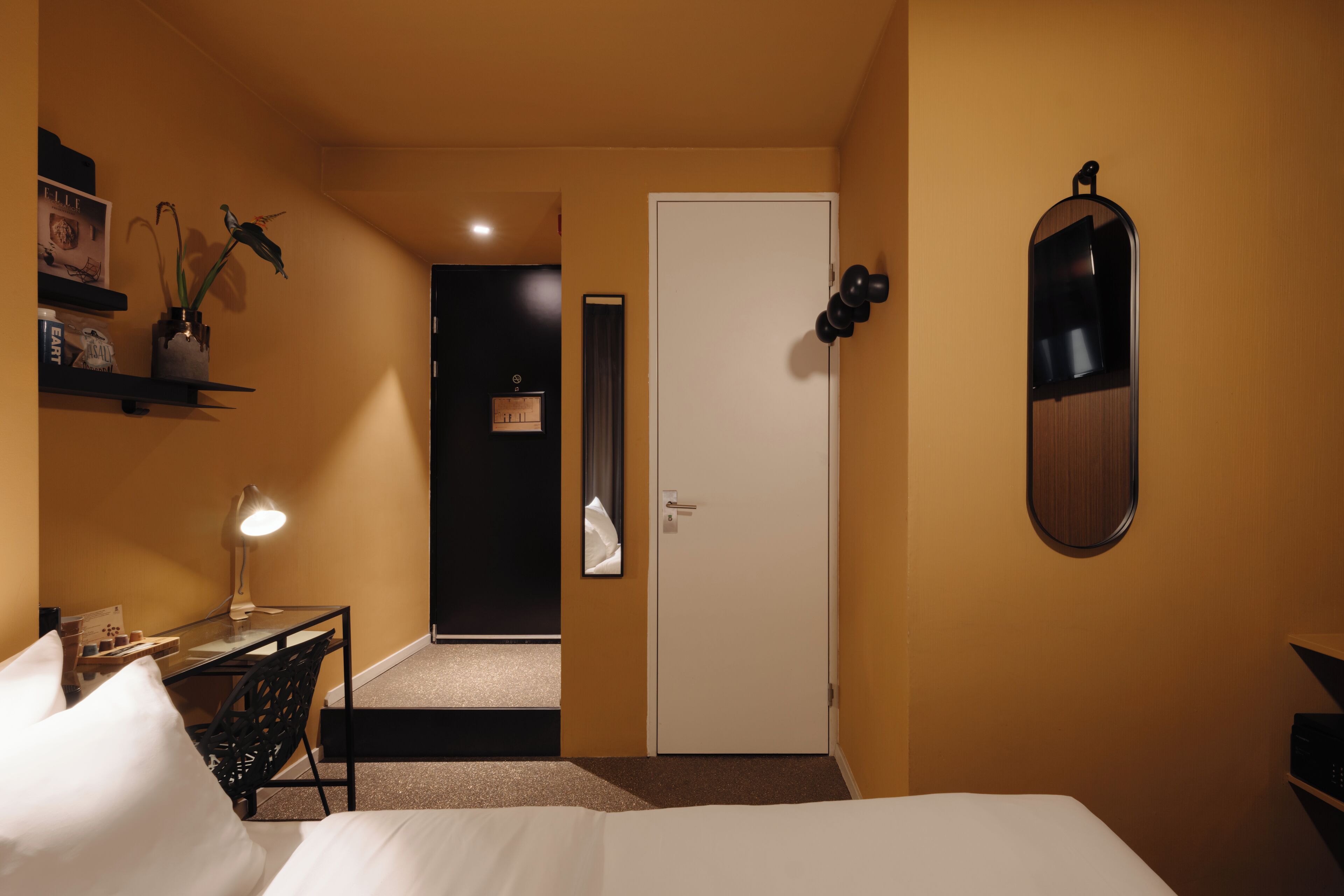 Quarto de hotel com cama, escrivaninha, cadeira, abajur e porta para um banheiro.