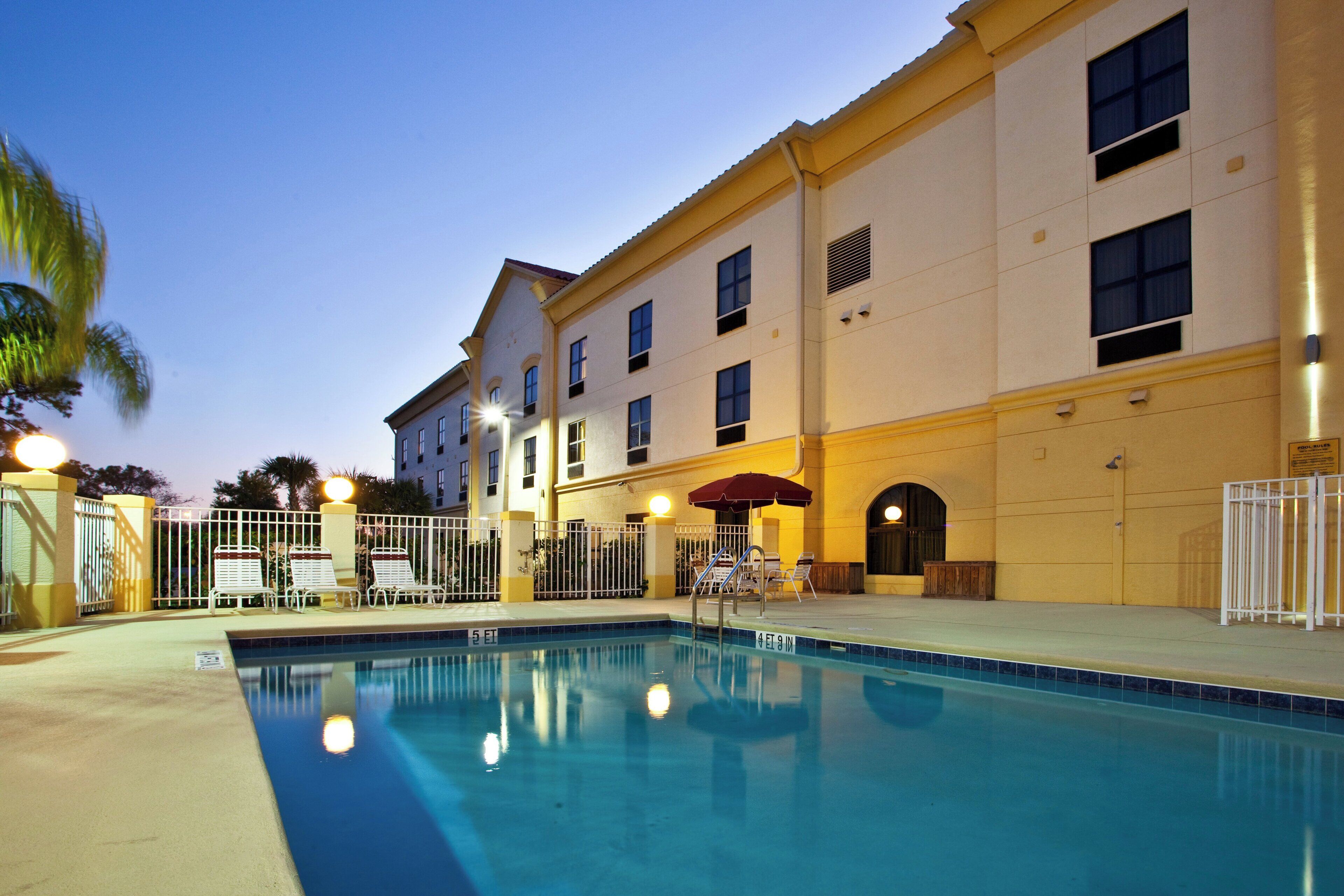 Área de piscina de hotel com piscina transparente, espreguiçadeiras e um edifício com várias janelas.
