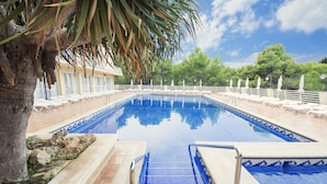 Uma piscina com bordas azulejadas, rodeada por espreguiçadeiras e guarda-sóis, com uma palmeira e um edifício ao fundo.