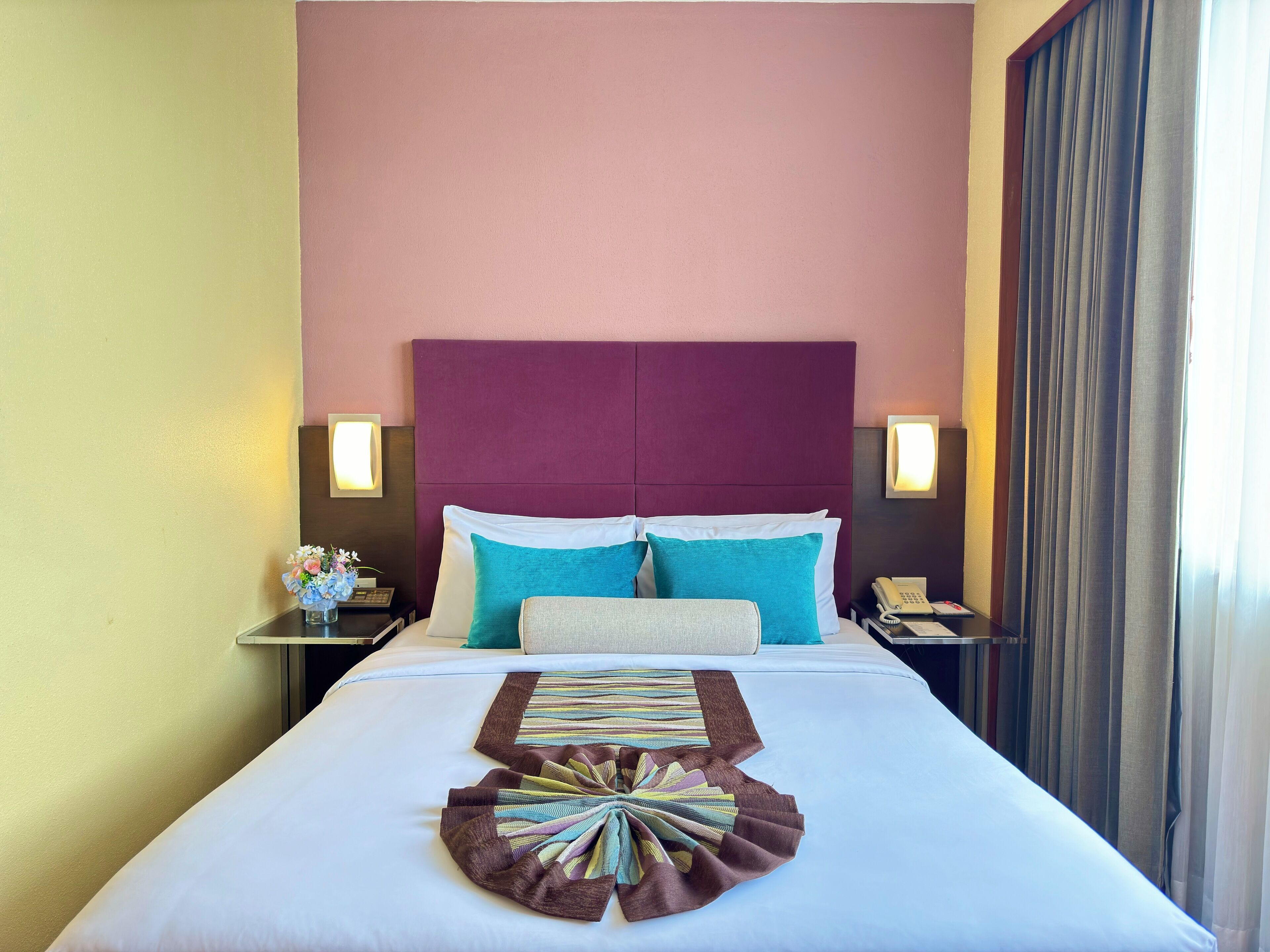 Quarto de hotel com uma cama grande, cabeceira, duas mesas de cabeceira com luminárias e uma janela com cortinas.