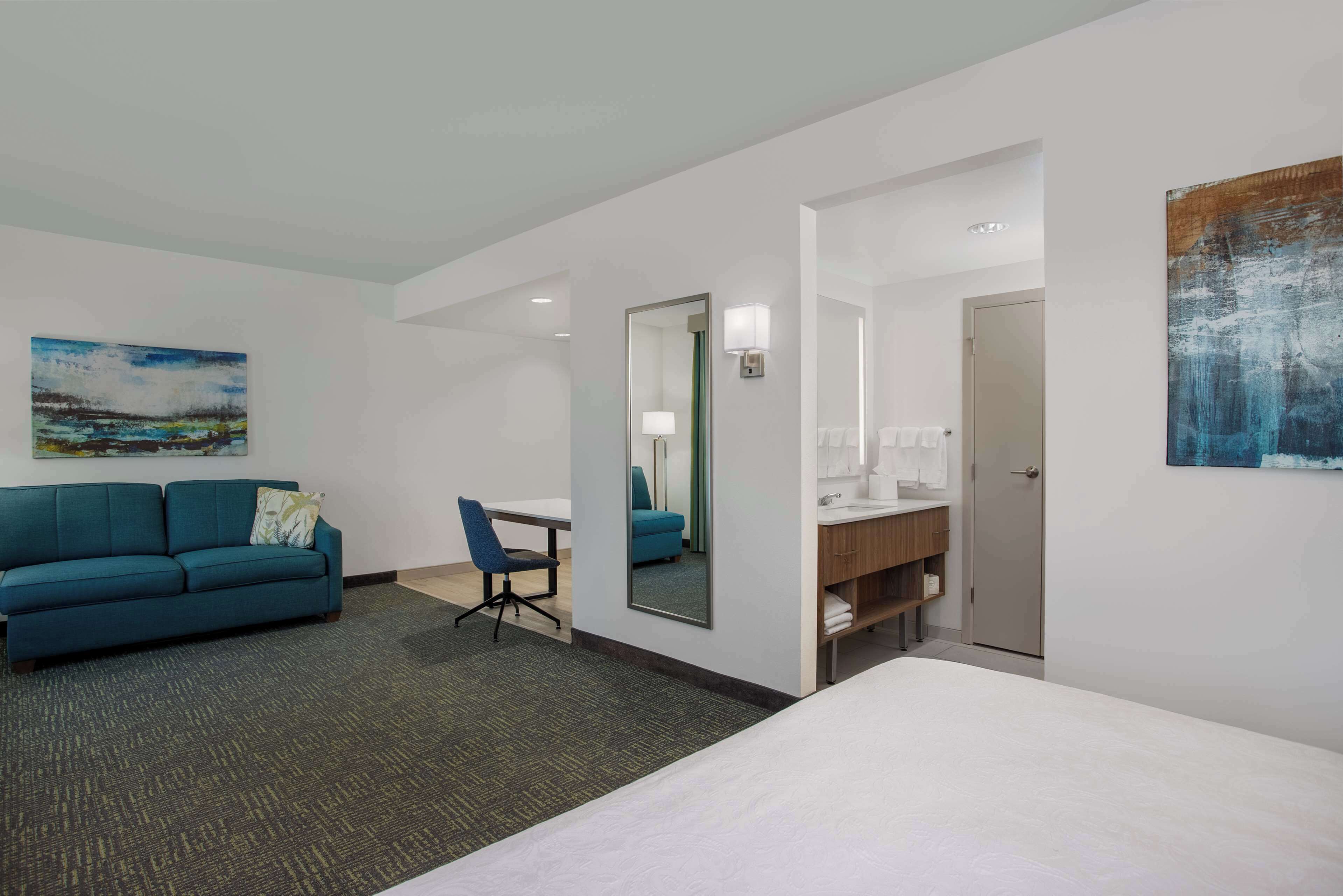 Quarto de hotel com uma cama, uma escrivaninha, um sofá azul, uma cadeira, um espelho e um quadro na parede.