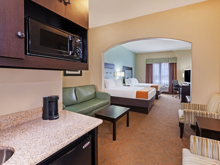 Quarto de hotel com cozinha compacta, sofá verde, cama com duas almofadas, escrivaninha e poltrona listrada.