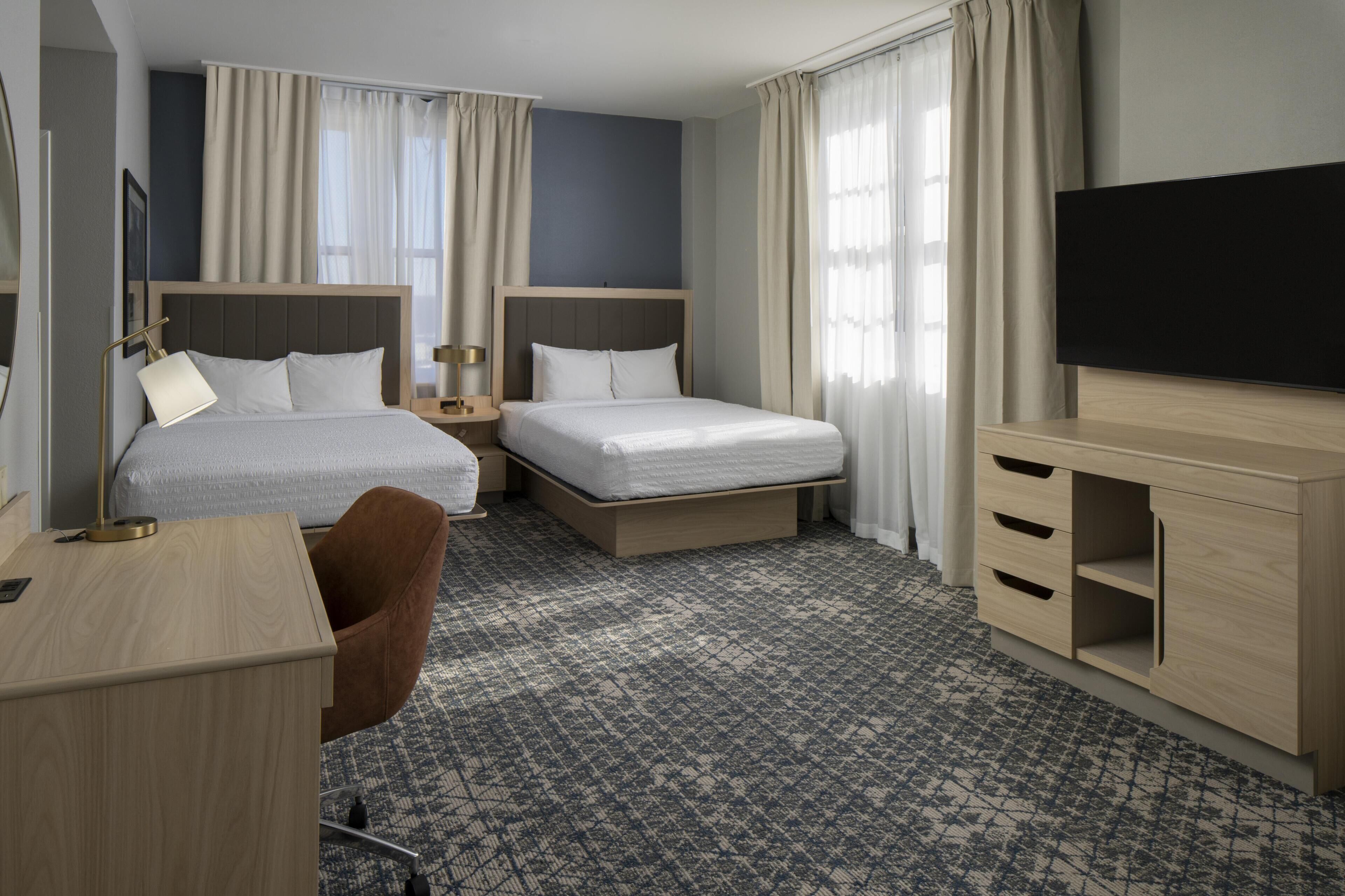 Quarto de hotel com duas camas, uma escrivaninha, uma cadeira, uma televisão e um guarda-roupa.