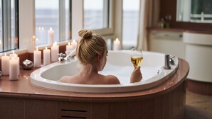 Mulher em banheira com velas e taça de vinho, grandes janelas e vista para a cidade.
