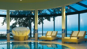 Uma piscina interior com paredes de vidro transparente, rodeada por cadeiras de vime e um grande sofá branco.