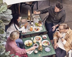 Um grupo de pessoas jantando em uma mesa ao ar livre, com diversos pratos, copos e um conjunto de chá.