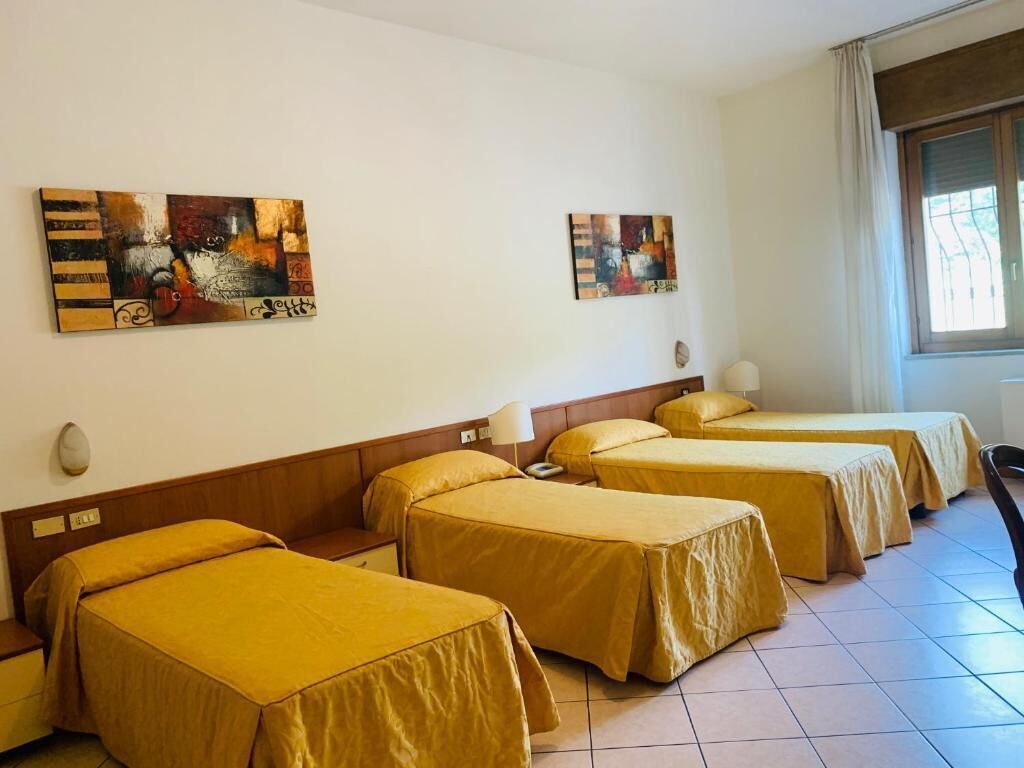 Quarto de hotel com quatro camas de solteiro, cabeceira de madeira, duas pinturas abstratas nas paredes, janela com cortinas e piso ladrilhado.
