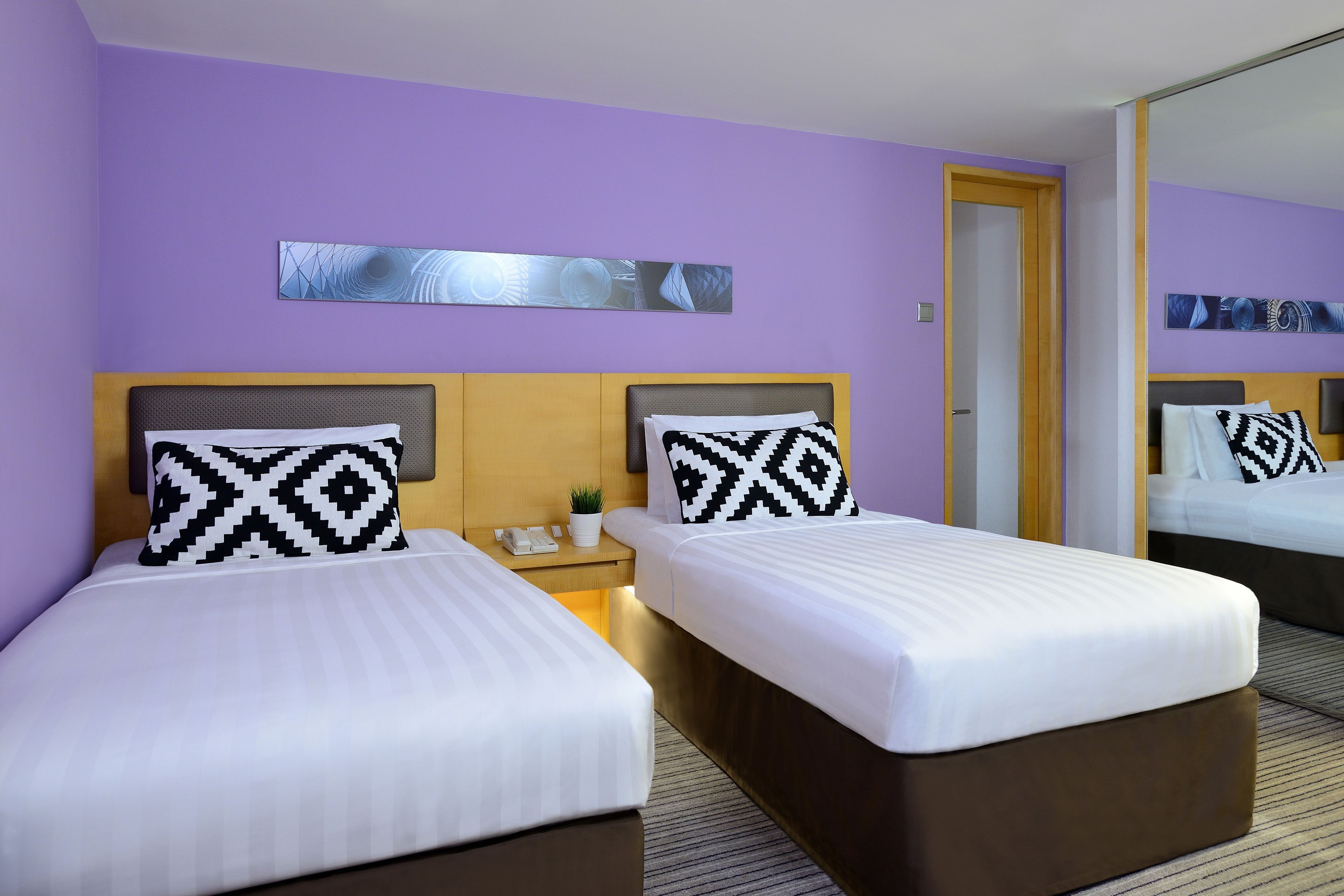 Quarto de hotel com duas camas, cada uma com almofada estampada, mesa de cabeceira com abajur e uma obra de arte na parede.