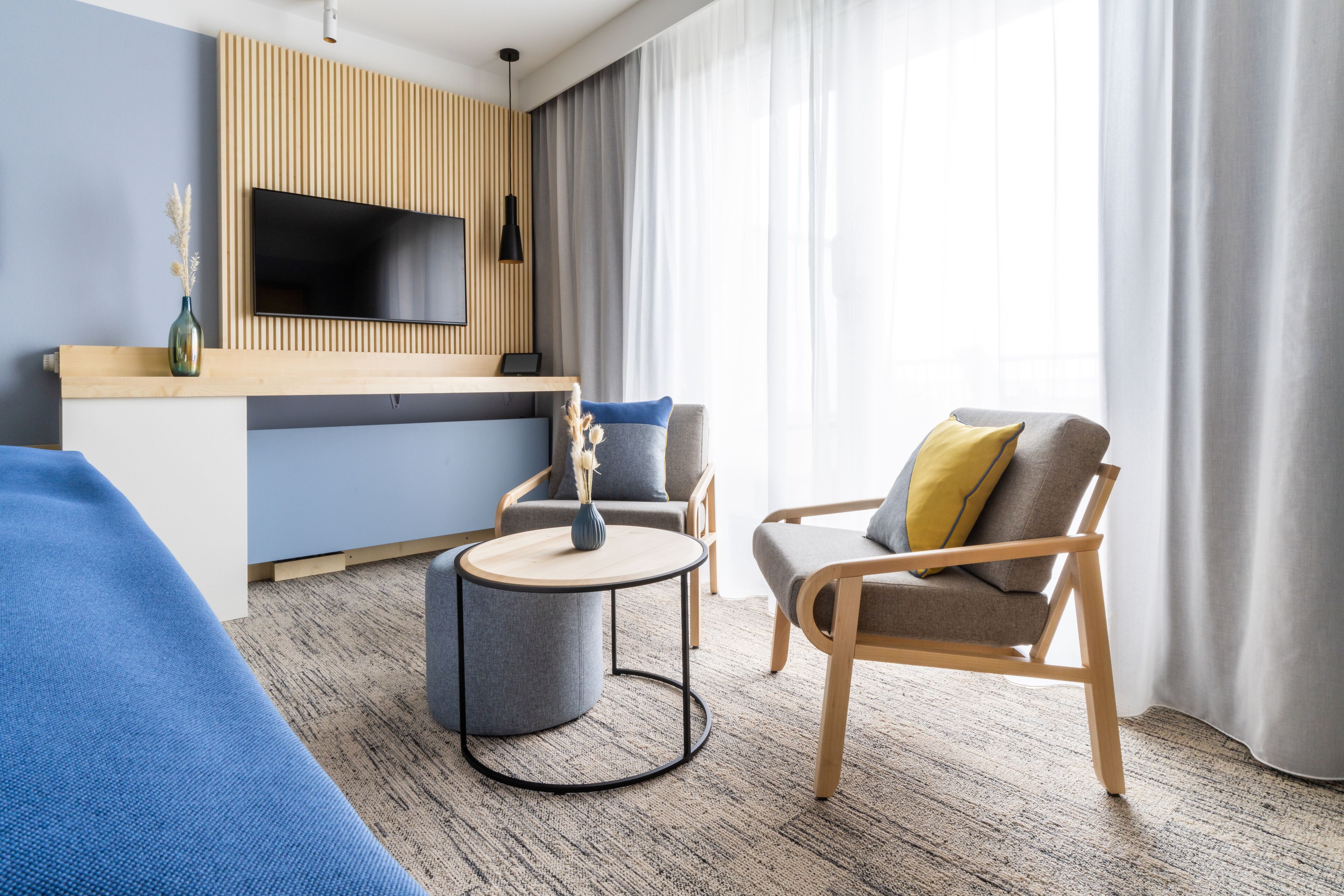 Quarto de hotel moderno com televisão de tela plana fixada em um painel de madeira, uma mesa de centro redonda, duas poltronas e um sofá.
