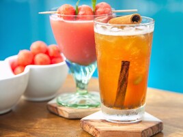 Duas bebidas em copos, uma delas com canela, e uma tigela de frutas ao fundo.