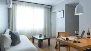Quarto de hotel moderno com um sofá branco, uma pequena mesa com um laptop e vinho, e uma mesa de jantar com cadeiras.