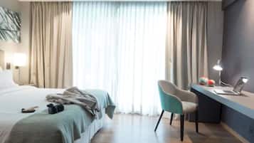 Quarto de hotel moderno com cama, escrivaninha, cadeira e janelas amplas com cortinas.