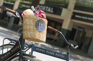 Uma bicicleta com uma cesta de vime contendo uma baguete e uma garrafa, estacionada em frente a uma placa com a inscrição 'Portal Plaza Suites'.