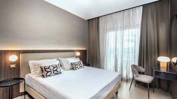 Quarto de hotel moderno com uma cama grande, uma mesa com cadeira e uma janela ampla com cortinas.