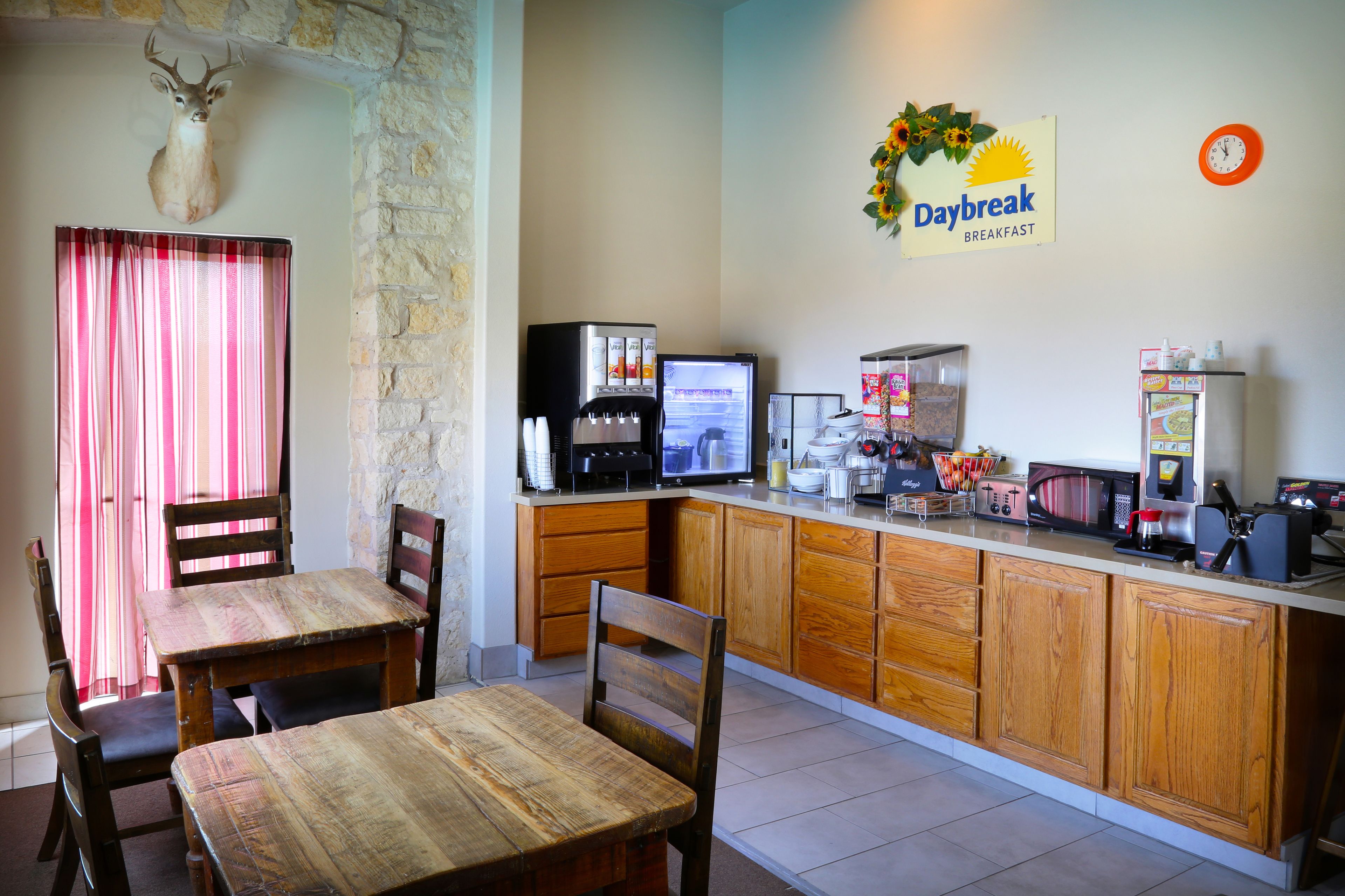 Uma área de café da manhã com móveis de madeira, uma geladeira, uma máquina de café e uma placa com a inscrição 'Daybreak Breakfast'.