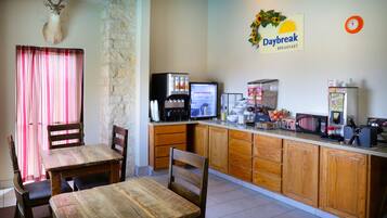 Uma área de café da manhã com móveis de madeira, uma geladeira, uma máquina de café e uma placa com a inscrição 'Daybreak Breakfast'.