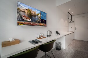 Quarto de hotel moderno com um balcão comprido branco, uma televisão na parede exibindo um canal de notícias, uma lixeira pequena e uma cadeira preta com almofada verde.
