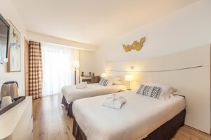 Quarto de hotel com duas camas, uma escrivaninha, uma cadeira e uma janela grande com cortinas.