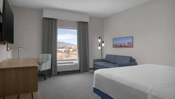 Quarto de hotel com cama, escrivaninha, cadeira, sofá, janela com cortinas, um quadro e uma TV de tela plana.