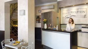 Uma área de recepção com balcão, bancada com um vaso de flores, quadro na parede e escada com corrimão decorativo.