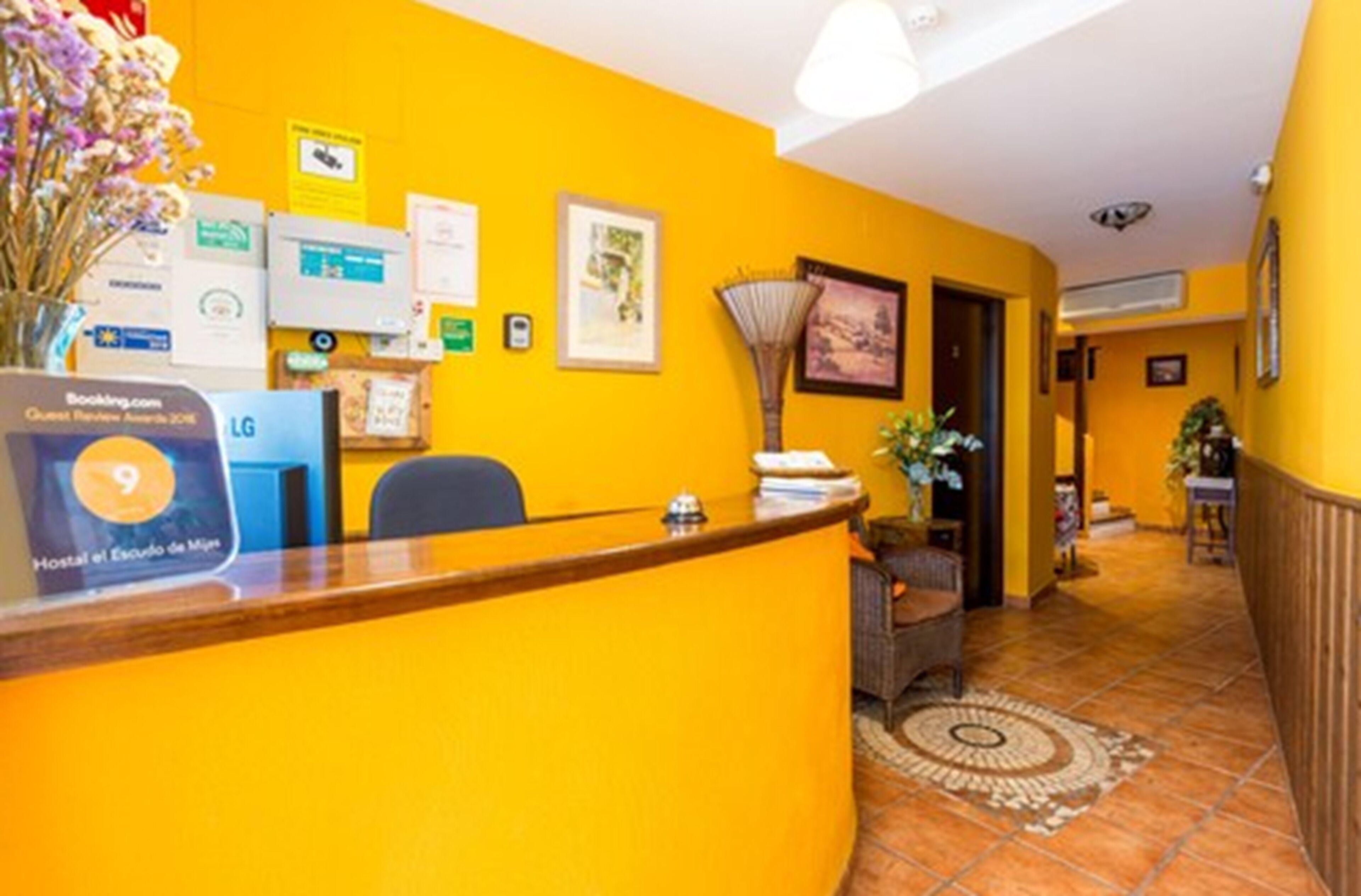 Uma área de recepção com parede amarela, balcão de recepção, vaso com flores, certificados emoldurados e piso de azulejos.