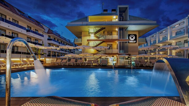 Uma área de piscina com espreguiçadeiras e guarda-sóis, um elemento aquático e um edifício com uma placa que diz 'JESSICA PARK'.