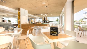 Restaurante moderno com mesas e cadeiras brancas, menu visível e vista da cidade através de grandes janelas.