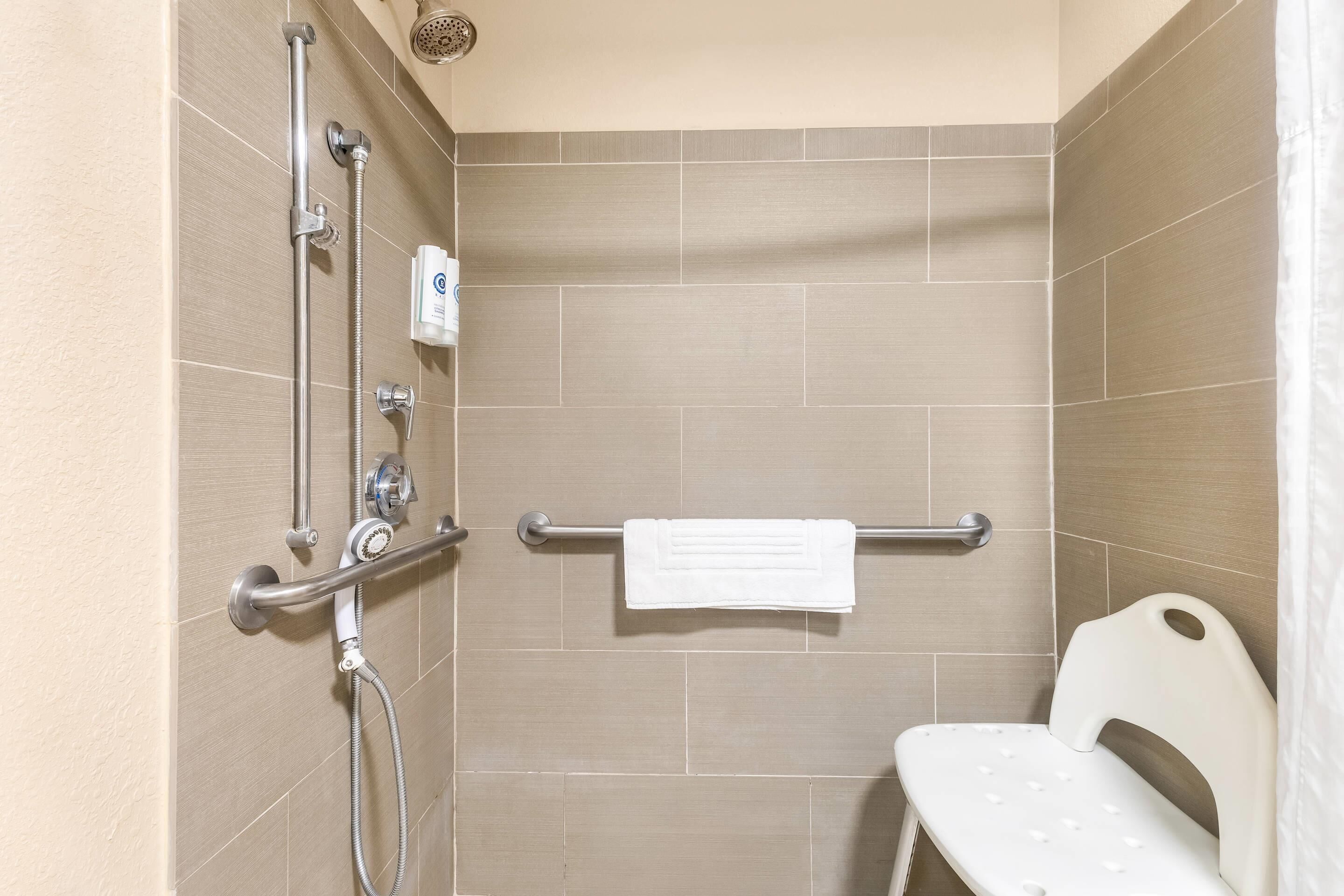 Um banheiro com azulejos bege, chuveiro com barra de mão, barra de apoio e porta-toalhas branco.