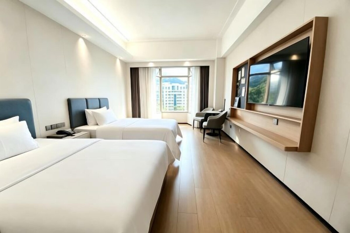 Quarto de hotel com duas camas, uma escrivaninha, uma cadeira, uma televisão e uma janela grande.