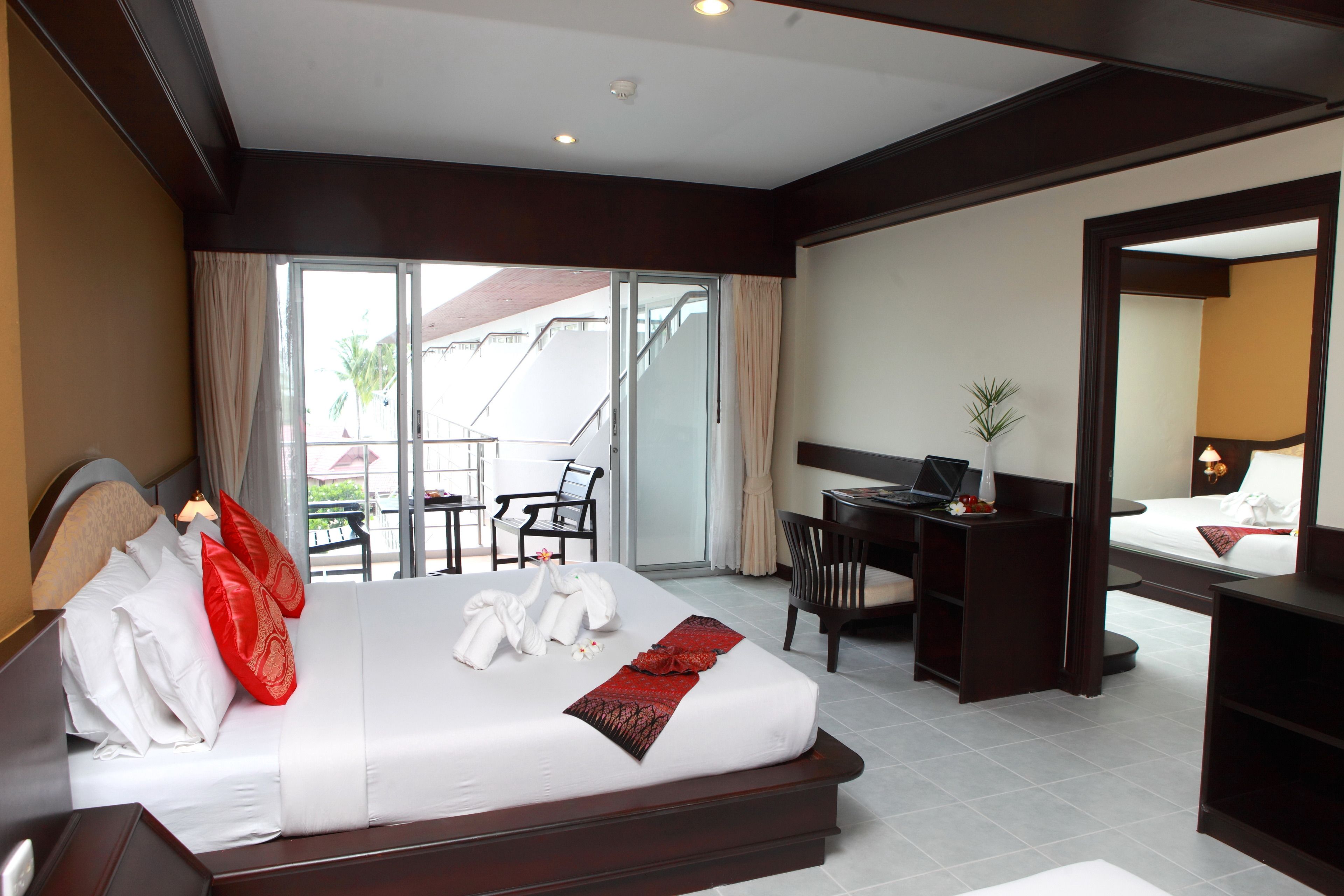 Quarto de hotel com uma cama grande, uma escrivaninha e uma cadeira. Tem uma varanda com vista para palmeiras e um edifício.