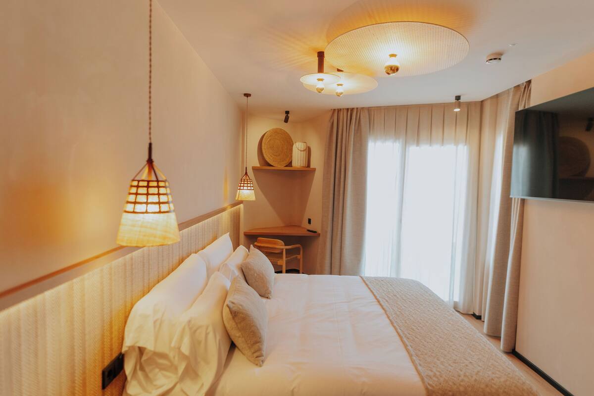 Quarto de hotel com cama, escrivaninha, cadeira, televisão e uma janela grande com cortinas.