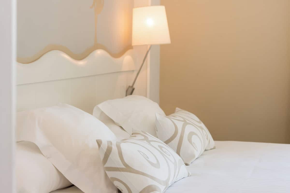 Uma cama bem arrumada com roupa de cama branca e almofadas estampadas, uma luminária fixada na parede e um desenho ondulado na cabeceira.