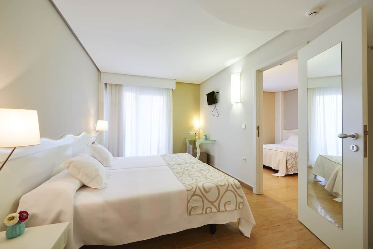 Quarto de hotel com duas camas, mesa de cabeceira com abajur, pequena mesa com luminária, TV fixada na parede e espelho na porta.