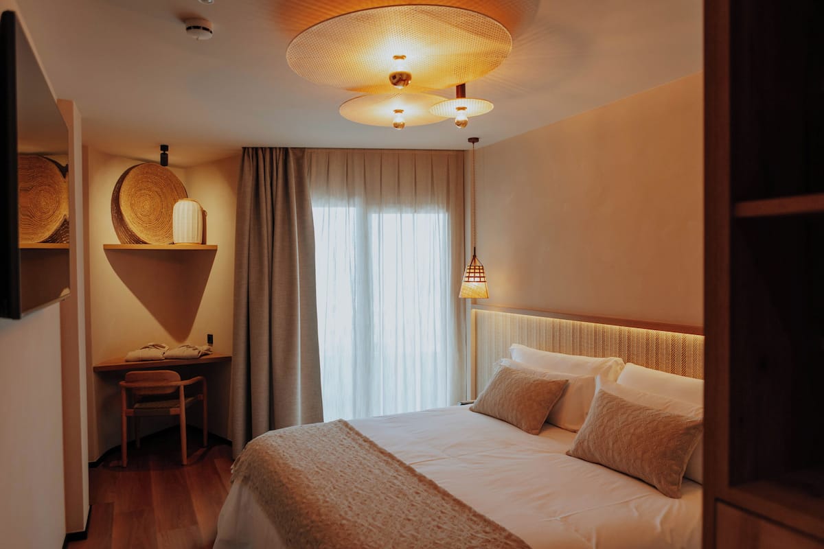 Quarto de hotel com uma cama, uma escrivaninha com cadeira, uma janela grande com cortinas e um lustre.