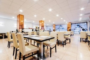 Ampla área de refeições do hotel, com várias mesas e cadeiras, um balcão de atendimento central e vista para a entrada do hotel.