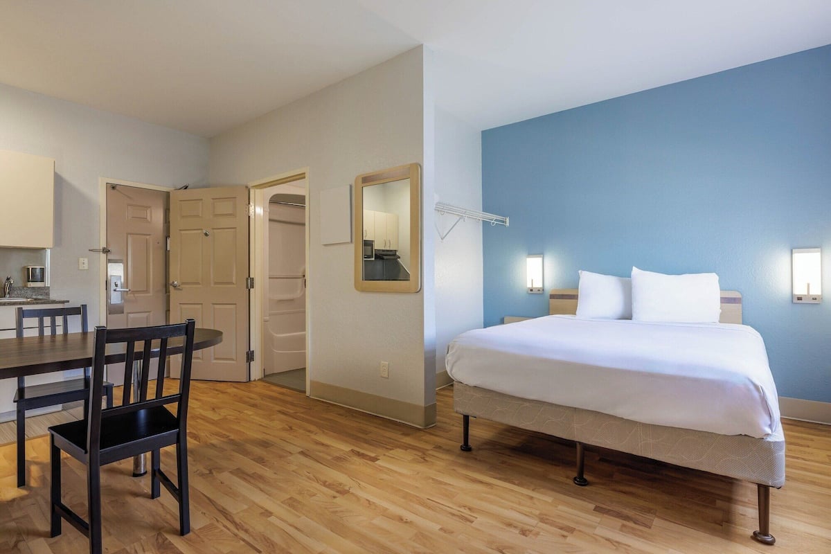 Quarto de hotel com cama, escrivaninha e cadeiras. Inclui banheiro com chuveiro e espelho.