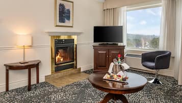 Quarto de hotel com lareira, televisão, mesa de jantar com uma cesta de frutas, sofá e vista para o exterior.