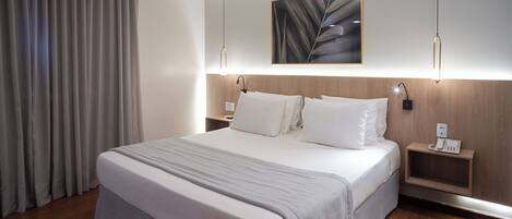 Quarto de hotel moderno com uma cama grande, cabeceira de madeira, mesas de cabeceira com luminárias e uma obra de arte na parede.