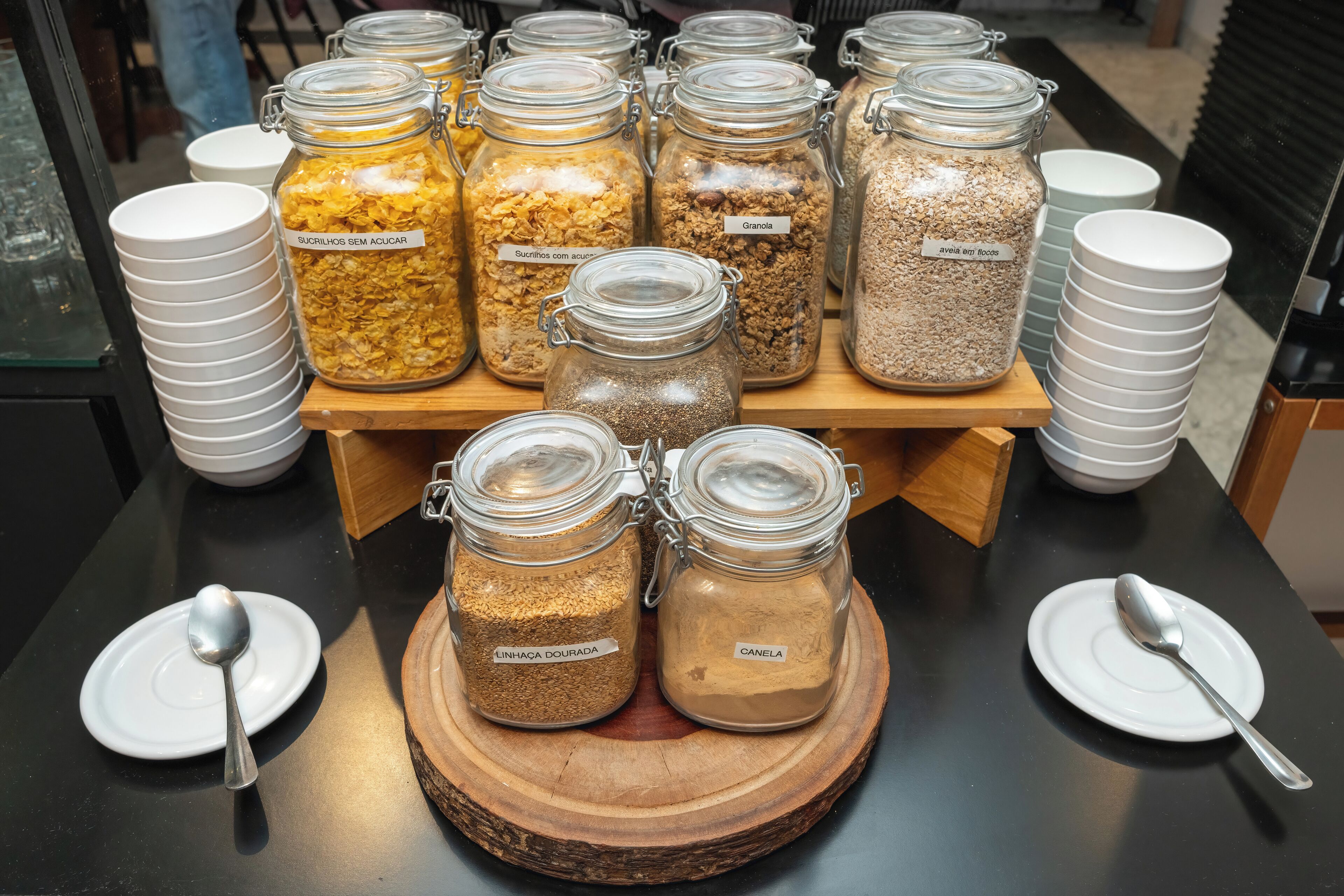 Mesa de buffet com diversos alimentos em frascos de vidro e pratos brancos com colheres.