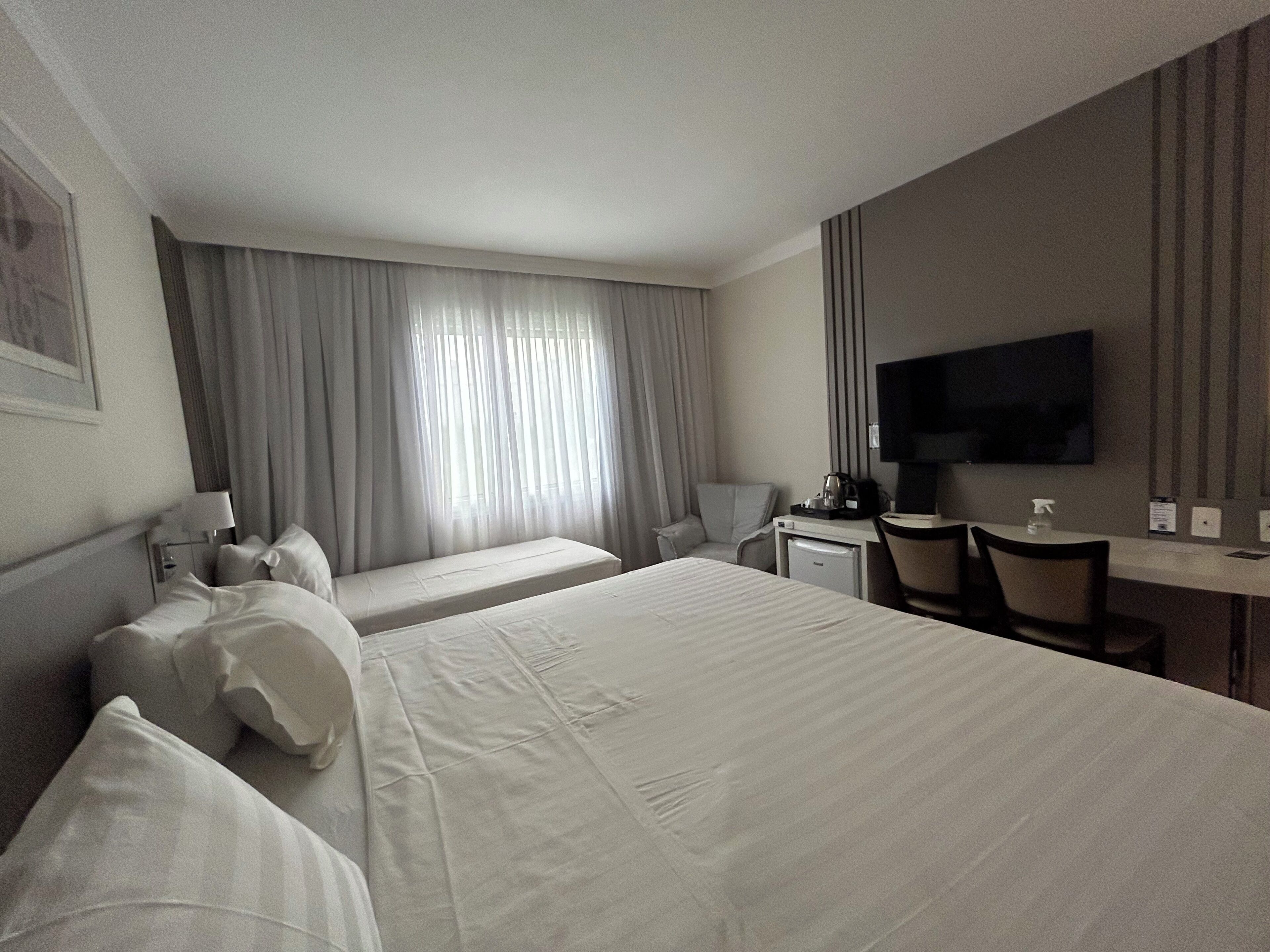 Quarto de hotel com duas camas, uma mesa, uma cadeira, uma televisão e uma janela com cortinas.