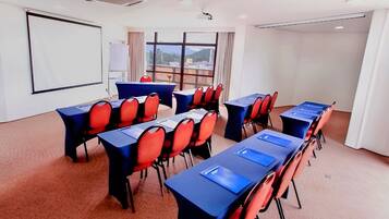 Uma sala de conferência com mesas, cadeiras, tela de projetor e vista da cidade através de grandes janelas.