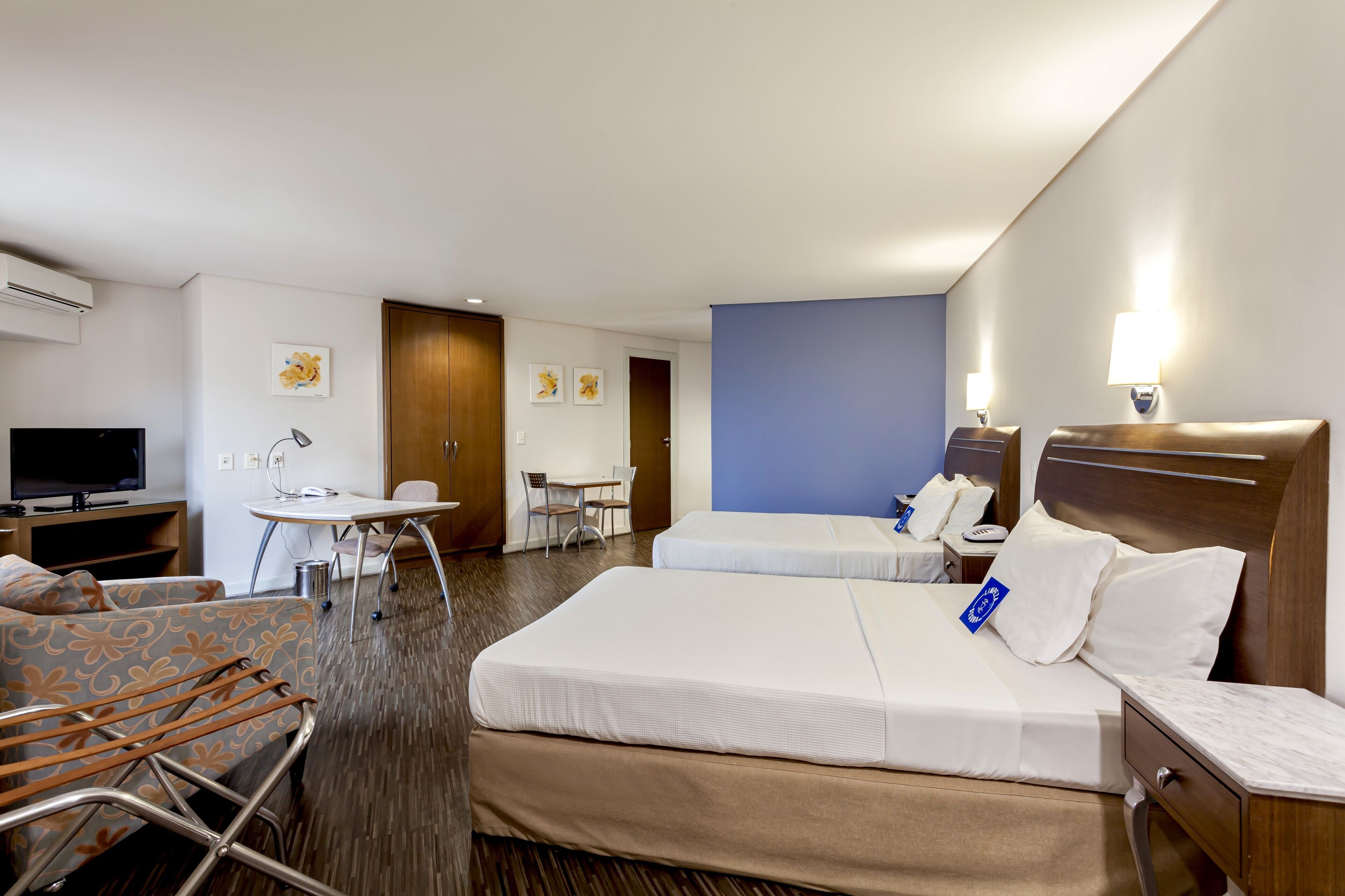 Quarto de hotel com duas camas, uma escrivaninha, uma cadeira, uma televisão e uma mesinha.