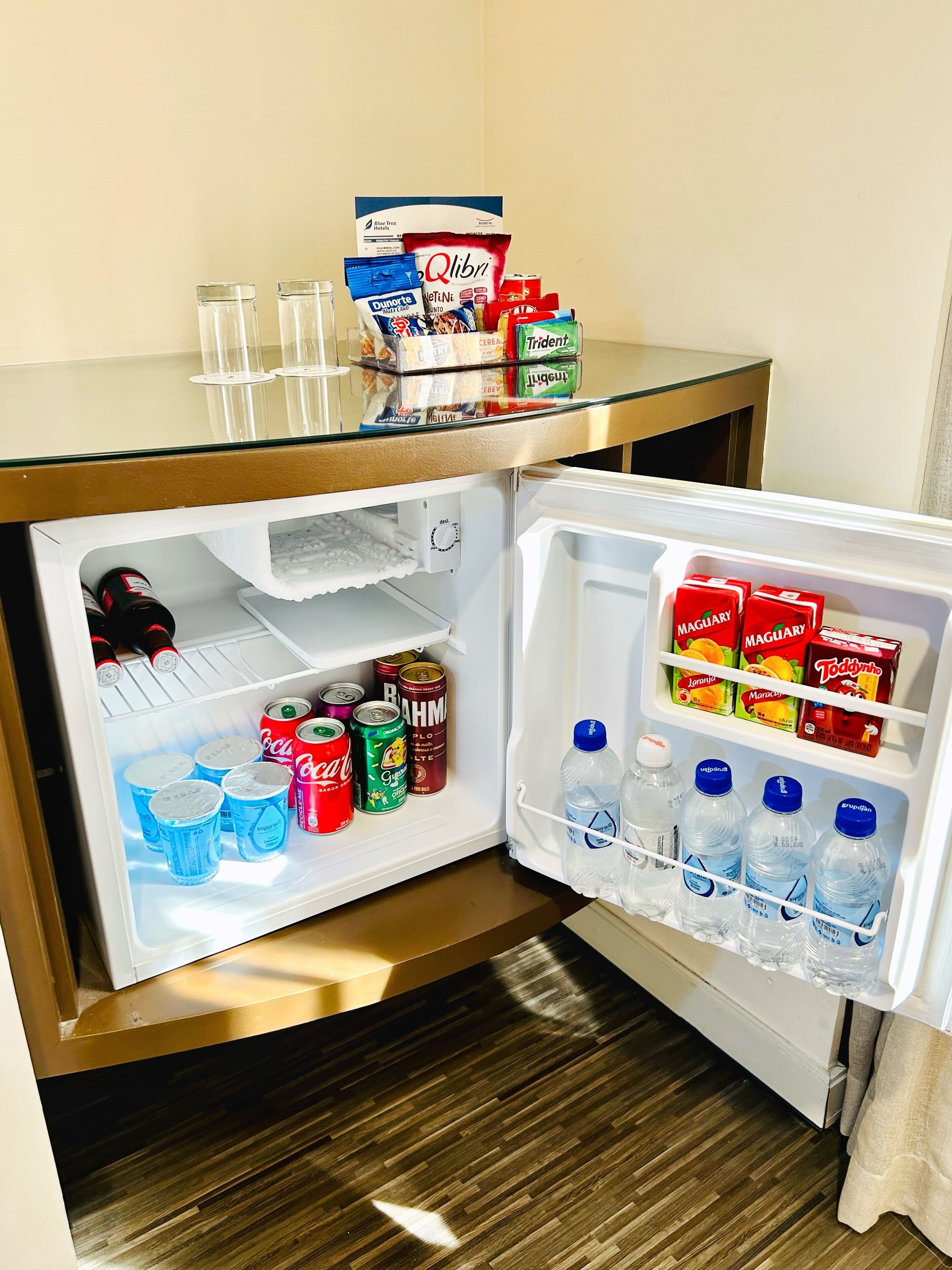 Mini-fridge com bebidas e petiscos, mesa de vidro com uma caixa azul e um pacote vermelho em cima, e piso de madeira.